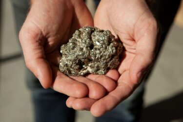A person holding a large, shiny, metallic-looking rock with both hands. The rock appears rough and glittery, resembling a lump of pyrite or "fool's gold.